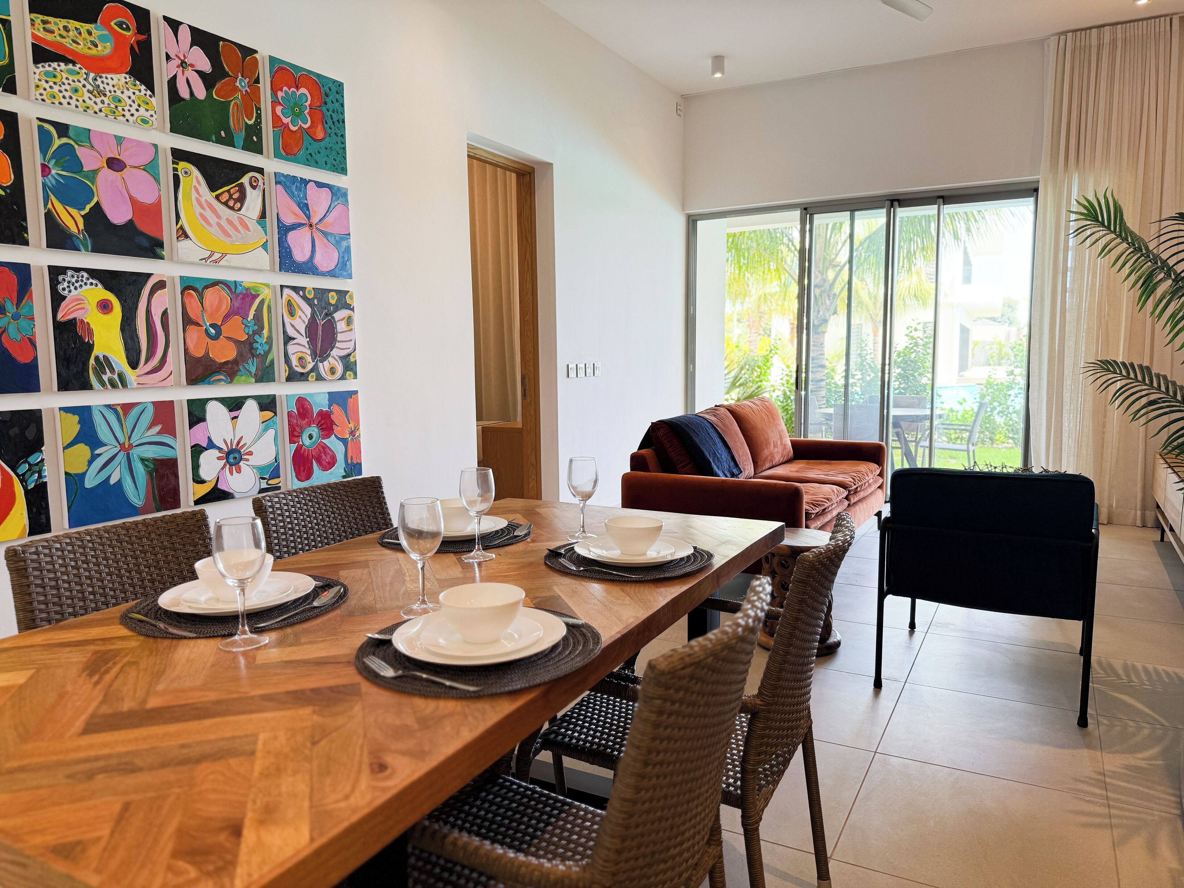 Uma área de refeições com uma mesa de madeira posta para quatro pessoas, rodeada por cadeiras de vime. Um sofá castanho e uma cadeira preta são visíveis ao fundo.