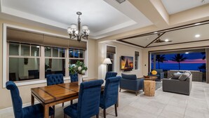 Uma sala de estar ampla com uma mesa de jantar de madeira, cadeiras estofadas em azul, um sofá cinza e vista para o oceano através de grandes janelas.