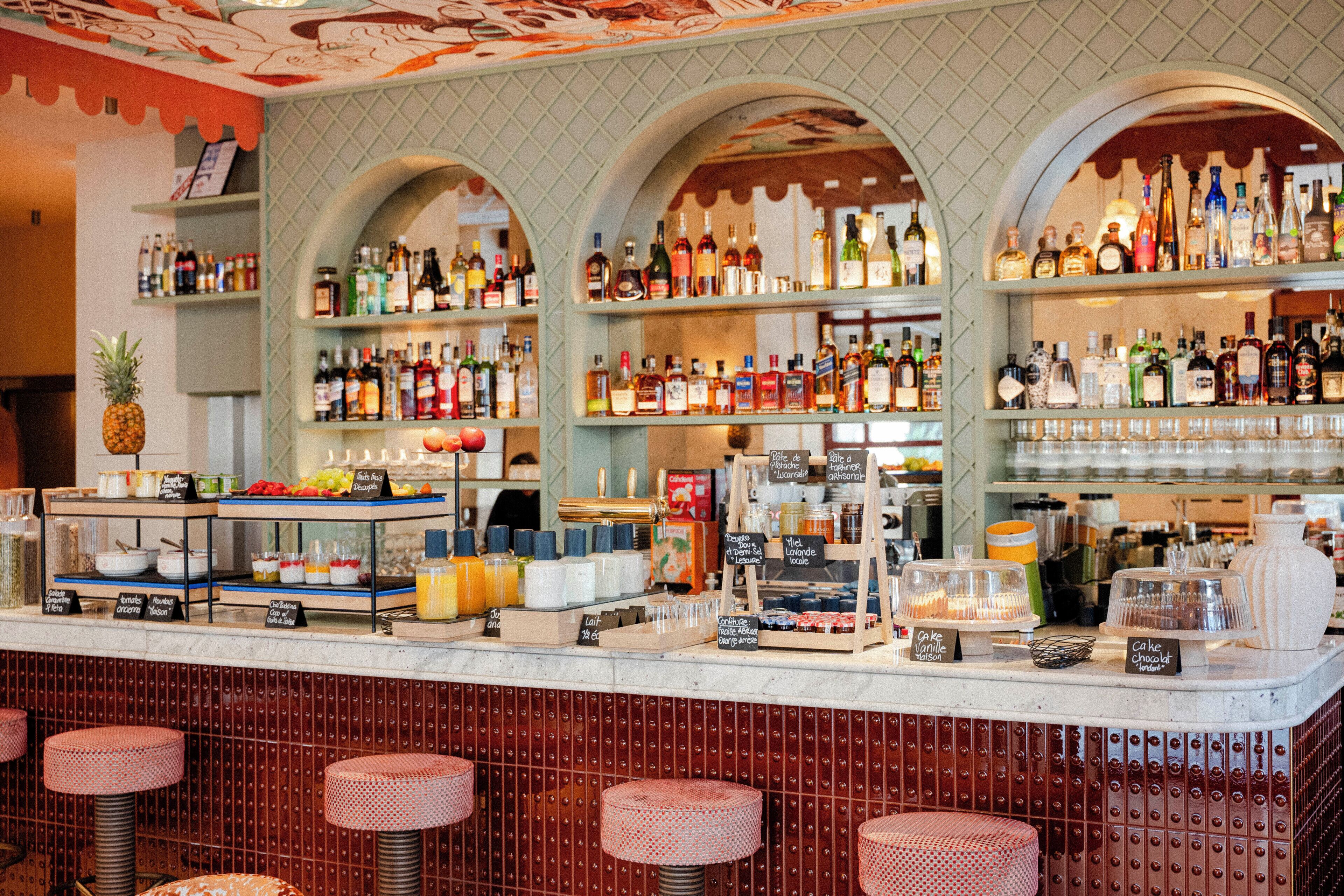 Bar com várias prateleiras repletas de garrafas, um expositor de frutas e um balcão com banquetas.
