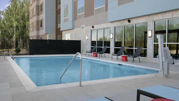Piscina com escada de metal, rodeada por espreguiçadeiras e um edifício com grandes janelas.