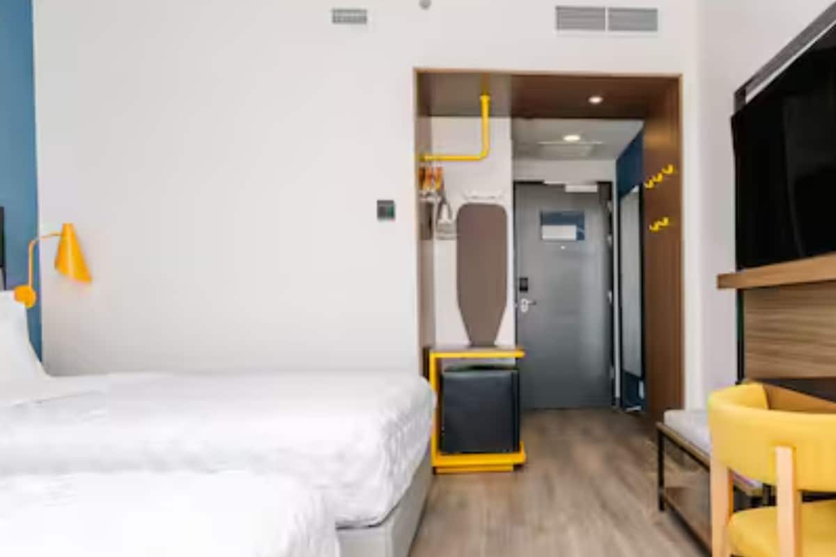 Quarto de hotel com cama, escrivaninha, cadeira, televisão e porta para o banheiro.
