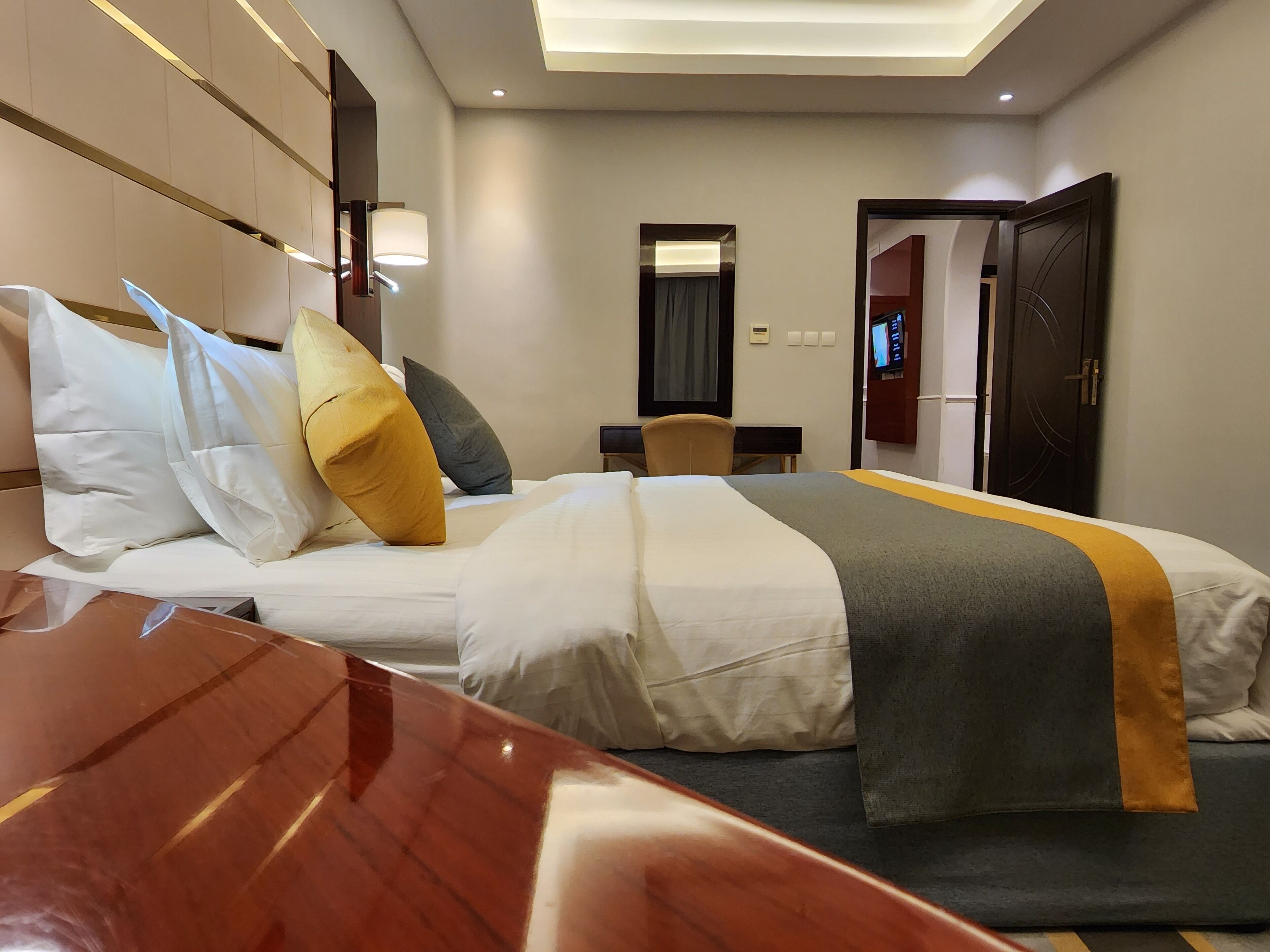 Quarto de hotel com uma cama bem arrumada, uma escrivaninha de madeira, uma cadeira, uma televisão e uma porta que dá para outro quarto.