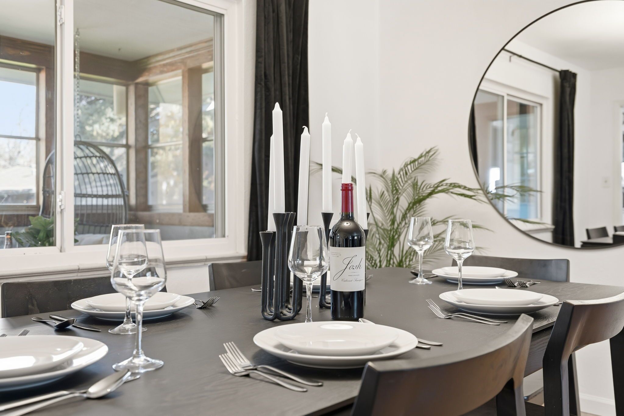 Uma mesa de jantar posta com pratos brancos, copos de vinho e uma garrafa de vinho. Há velas e um espelho sobre a mesa.