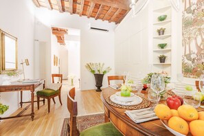 Uma sala de jantar com uma mesa de madeira posta para uma refeição, cercada por cadeiras, um vaso com flores e uma fruteira.