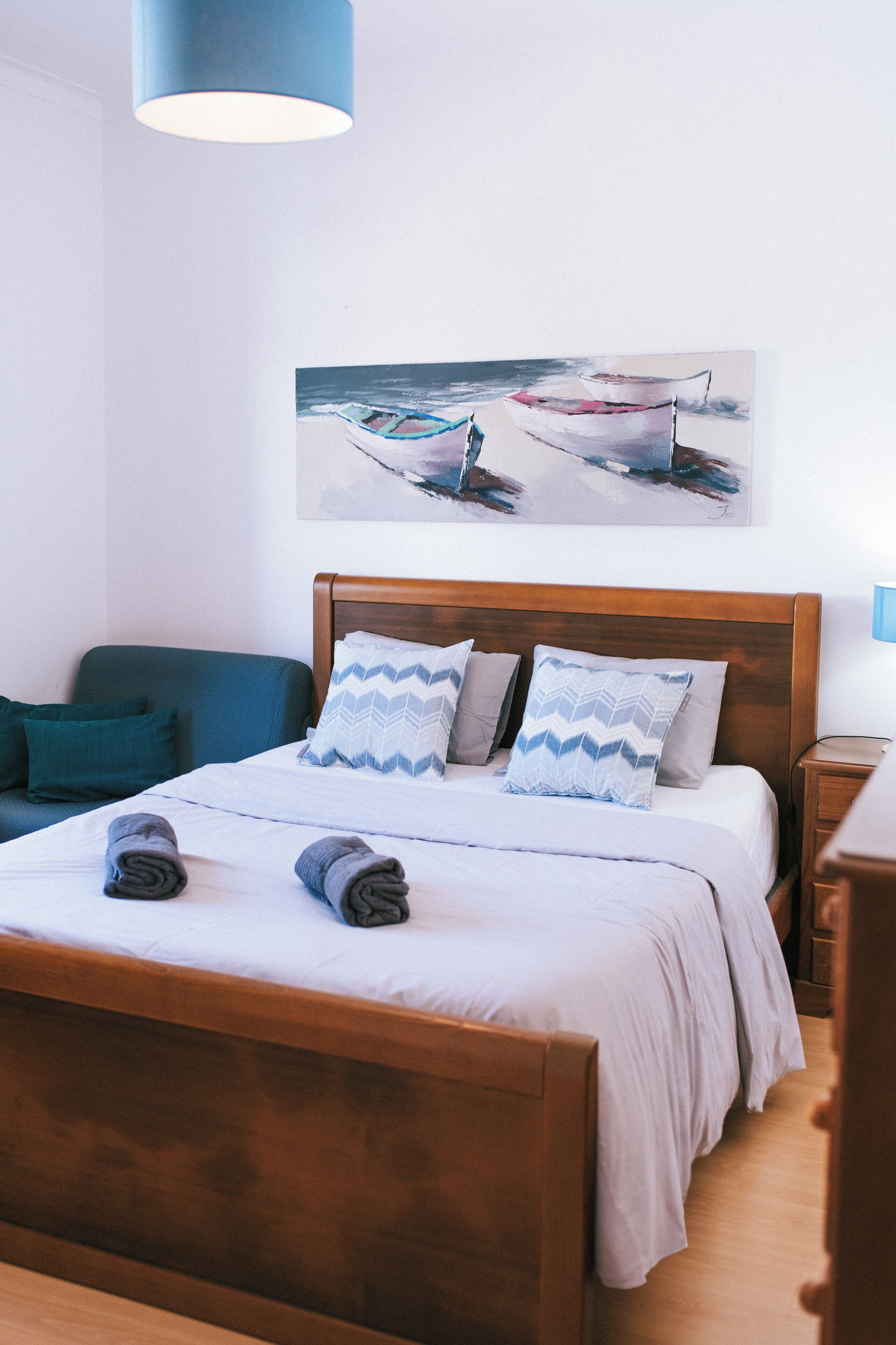 Uma cama bem arrumada com cabeceira de madeira, um abajur azul pendurado e um quadro com barcos na parede.