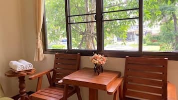 Quarto com mesa e cadeiras de madeira, janela com vista para vegetação e um vaso com flores.