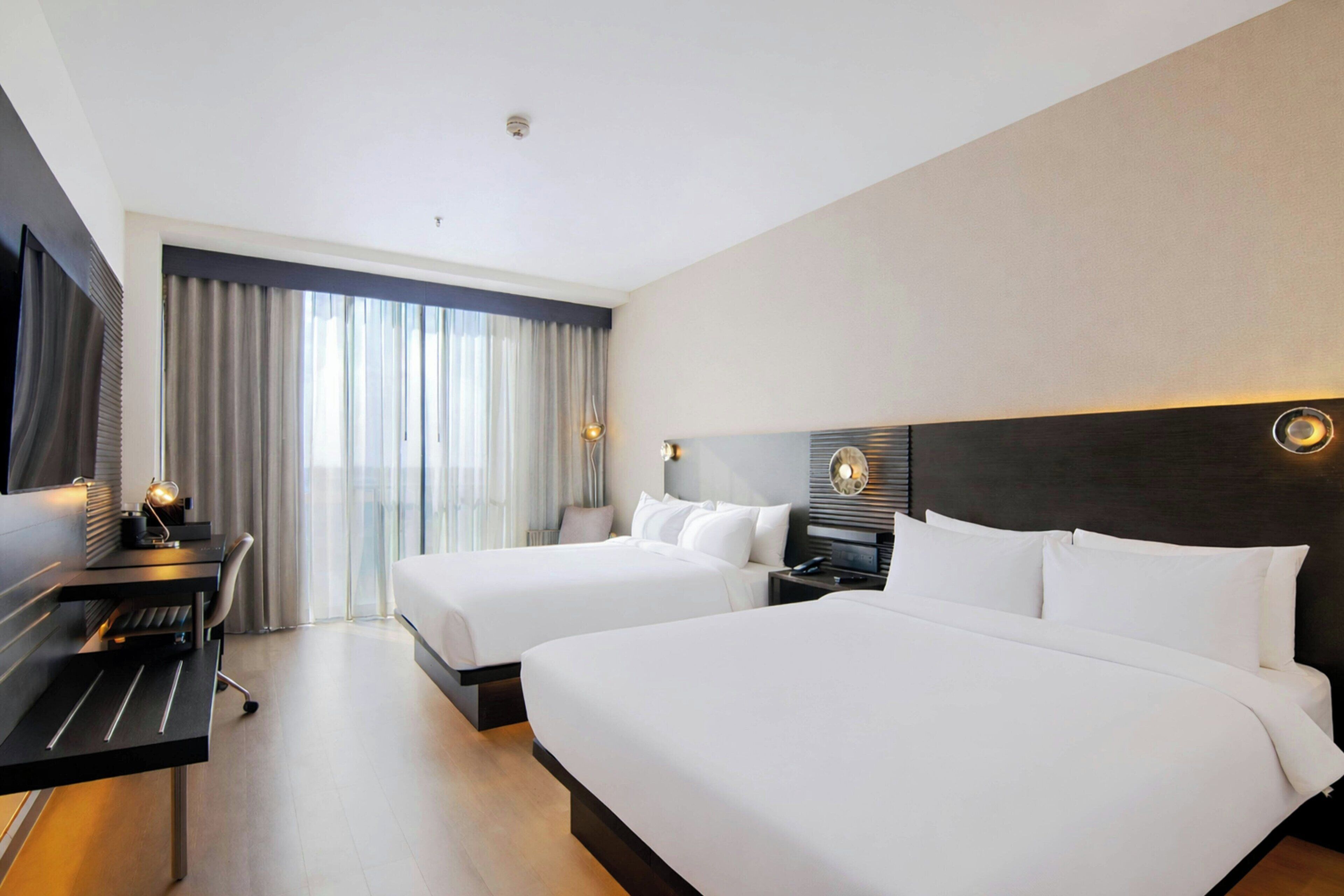 Quarto de hotel com duas camas, uma mesa, uma televisão e uma janela grande com cortinas.
