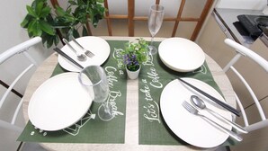 Uma mesa de jantar posta com pratos brancos, talheres e um copo de água, com um jogo americano verde e uma pequena planta em vaso.