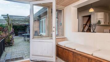 Quarto com um banco de madeira, porta que dá para área externa e cozinha visível através de uma porta aberta.