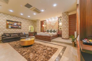 Quarto de hotel moderno com sofá, mesa de centro redonda, cama com almofadas decorativas e uma grande obra de arte na parede.