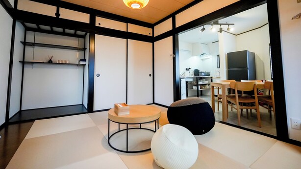 Um quarto japonês tradicional com tatames, uma mesa redonda com almofada, luminária pendente e portas deslizantes que dão para uma cozinha.