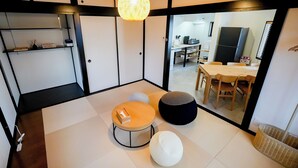 Um interior moderno com piso de tatame, uma mesa redonda de madeira, uma caixa pequena, um banquinho preto e uma luminária pendente trançada.