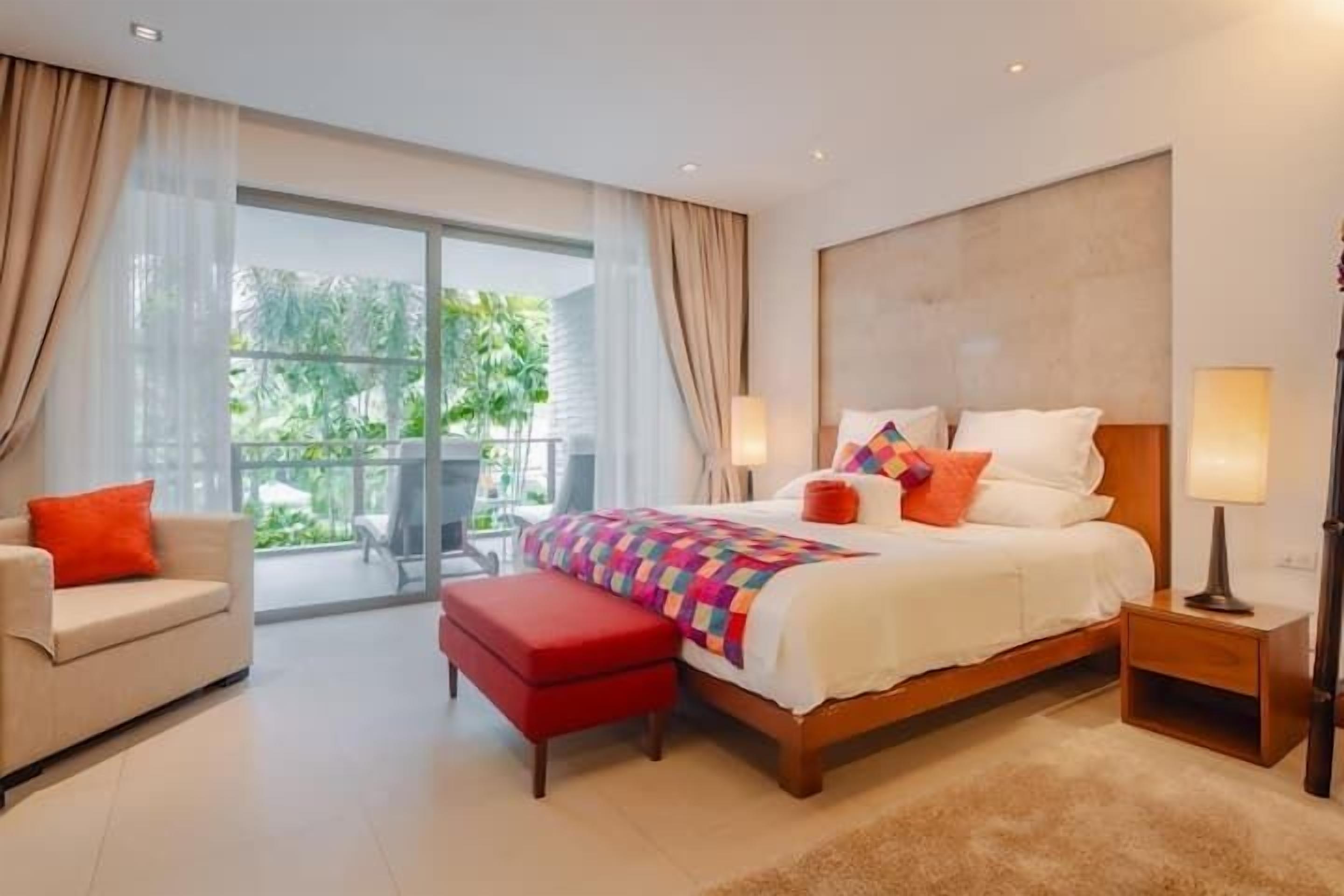 Quarto de hotel moderno com cama grande, um banco vermelho, sofá e varanda com vista para o verde.