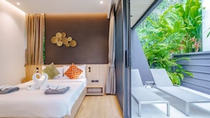 Quarto de hotel moderno com cama, uma mesinha de cabeceira, um objeto decorativo na parede e uma varanda com cadeiras de estar e vegetação.