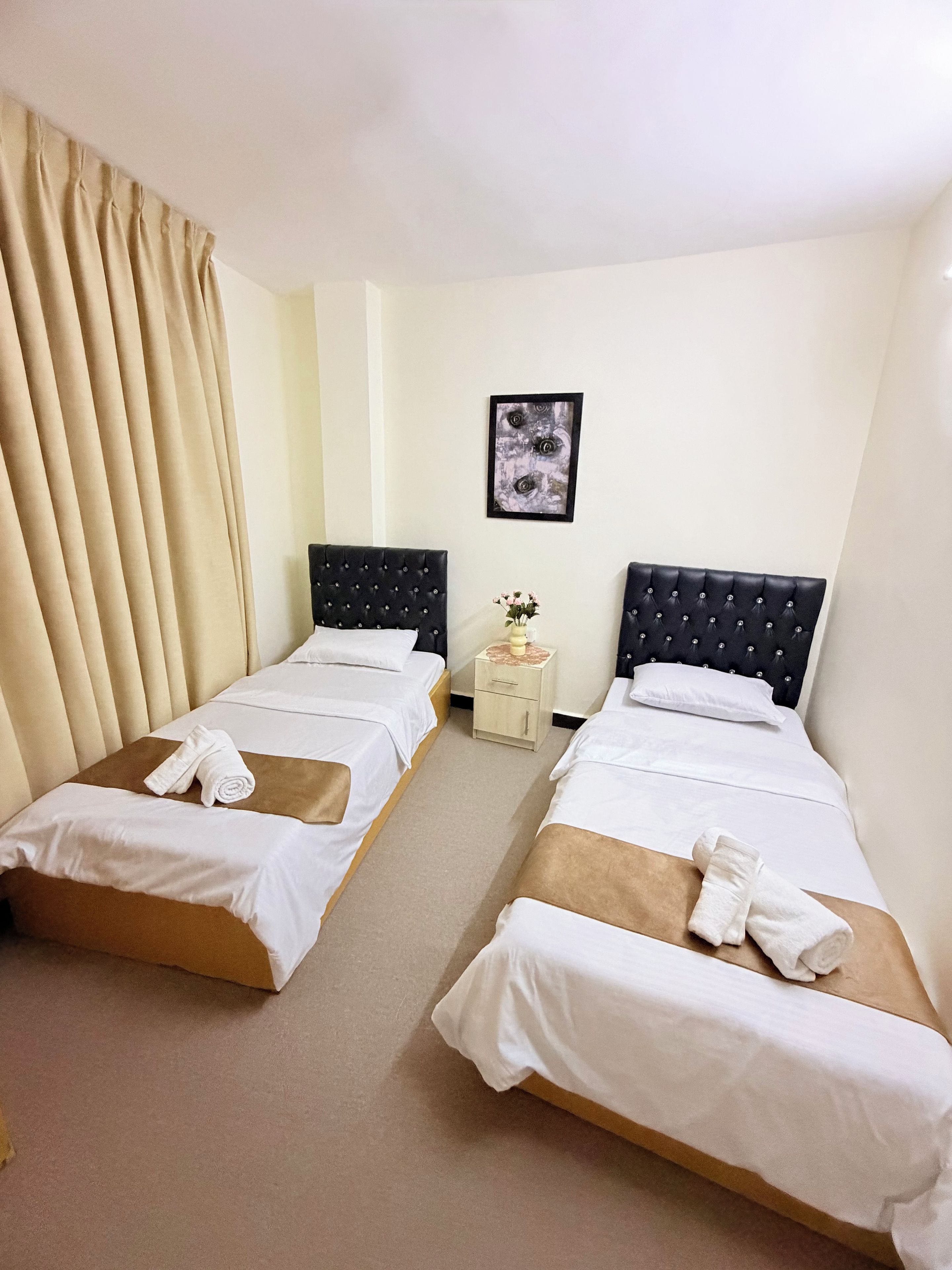 Quarto de hotel com duas camas, cortinas bege, uma pequena mesa de cabeceira com um vaso e um quadro pendurado na parede.