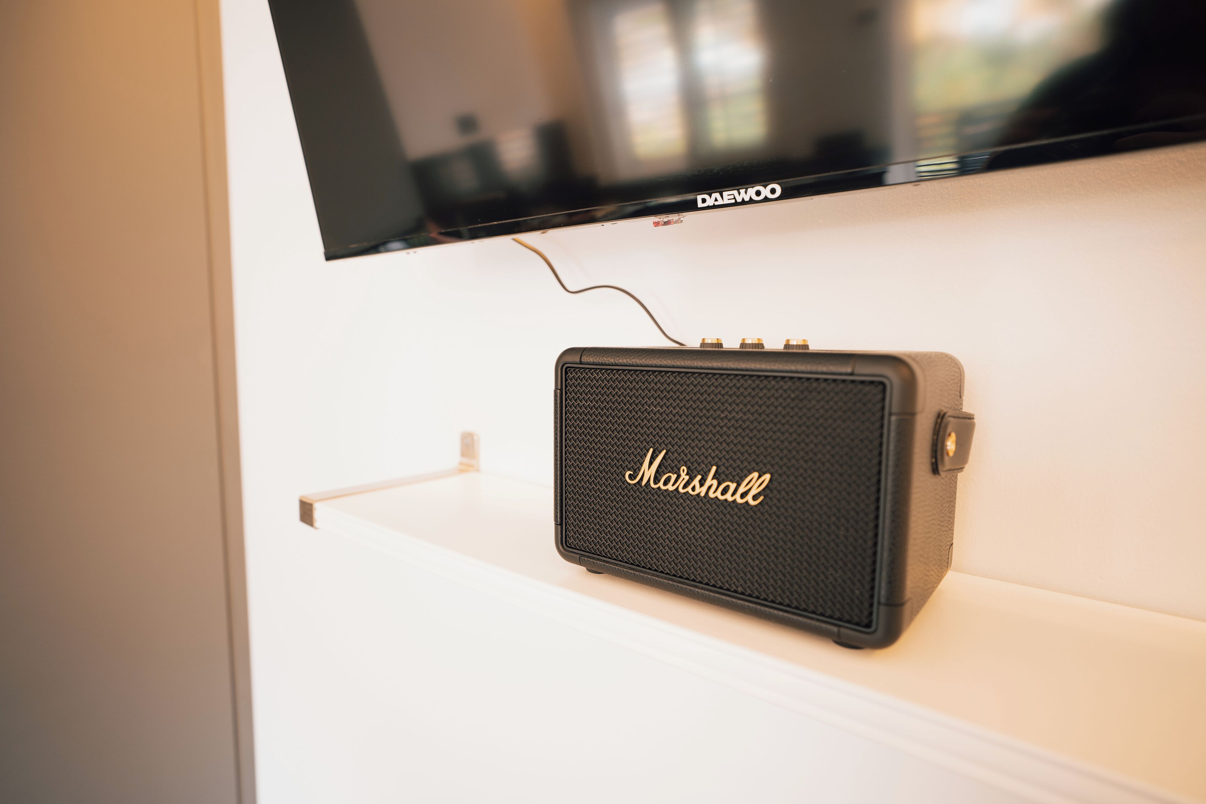 Uma caixa de som Marshall fixada em uma parede branca, com uma televisão acima.