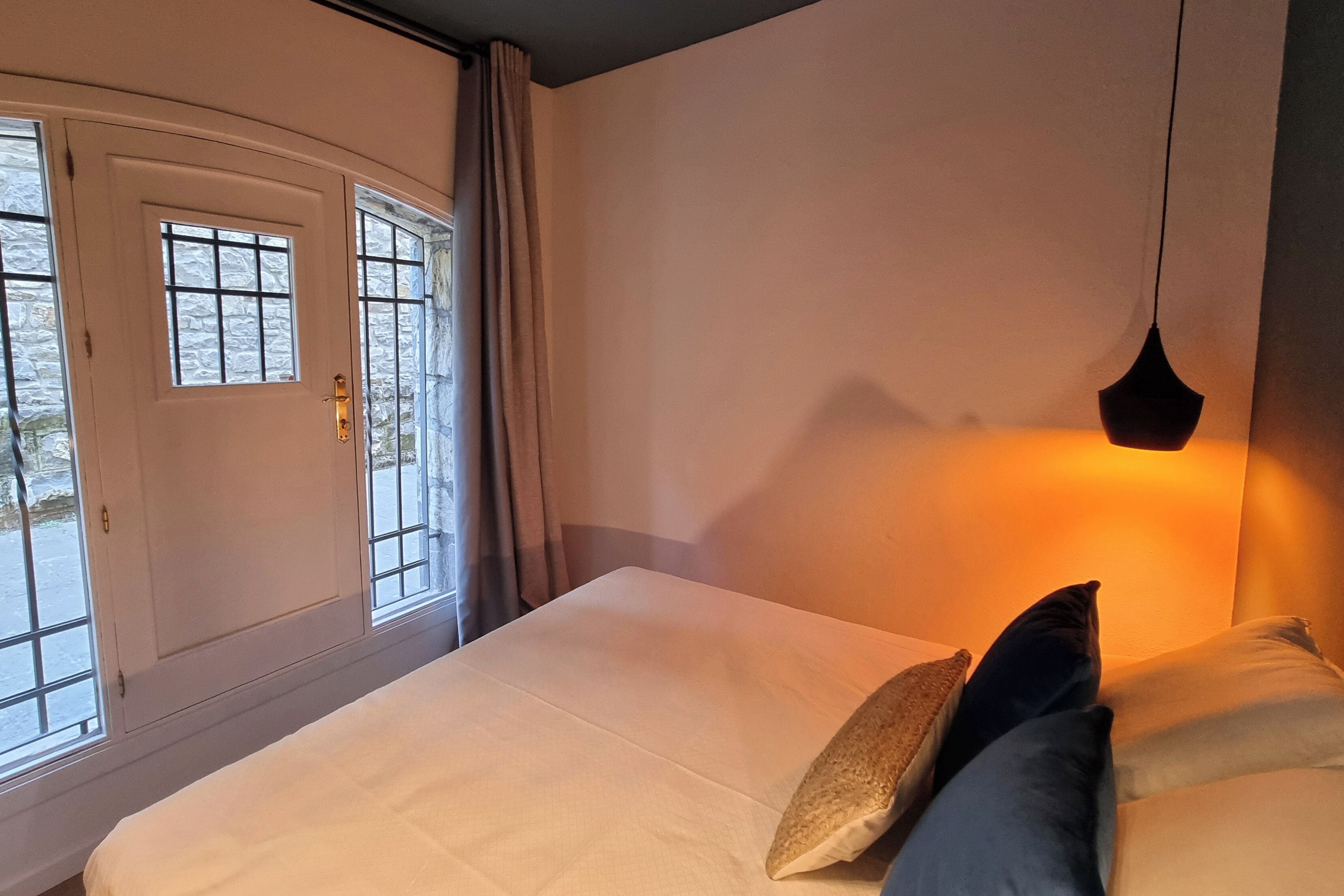 Quarto com cama, luminária pendente e porta com janelas.