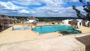 Uma grande piscina ao ar livre com várias áreas, rodeada por espreguiçadeiras e guarda-sóis, com edifícios de telhado de palha e um céu claro ao fundo.