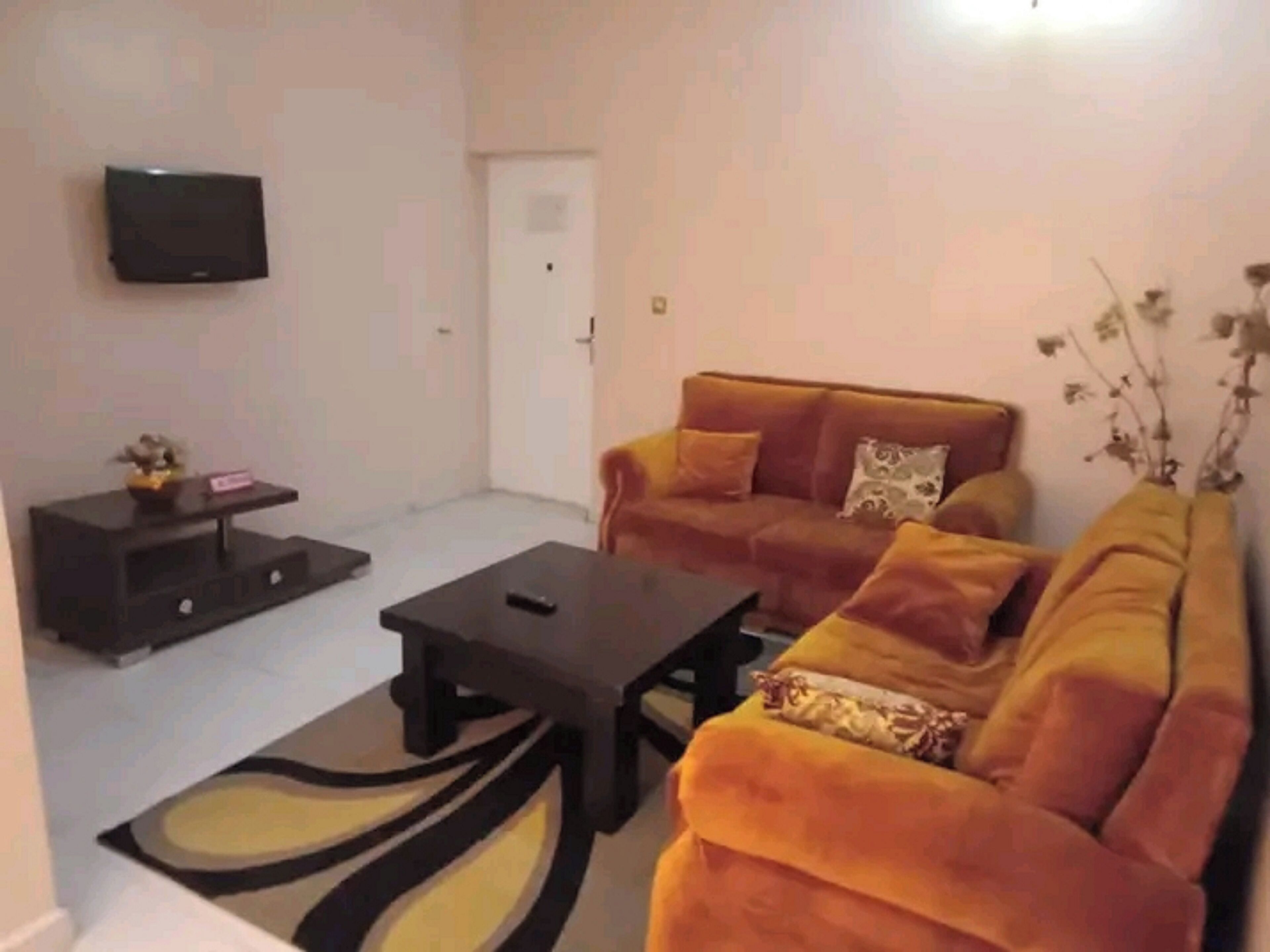 Uma sala de estar com sofá, mesa de centro, televisão fixada na parede e uma pequena mesa com objetos decorativos.