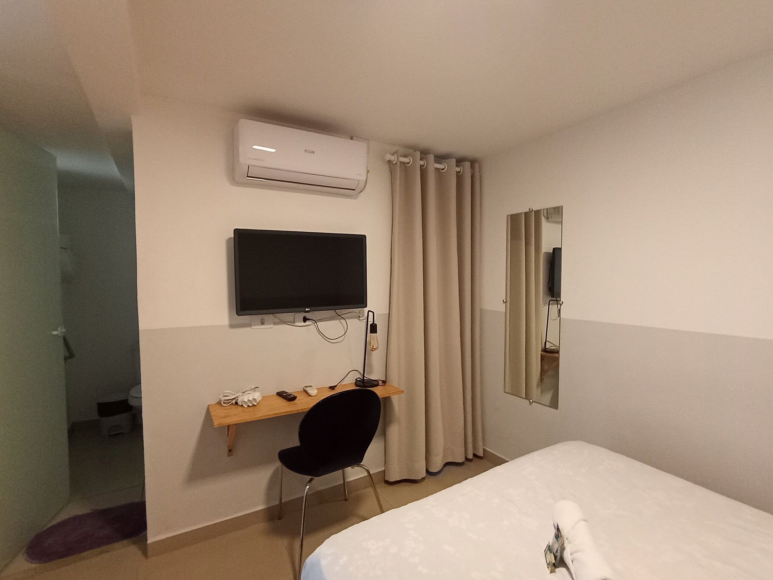 Quarto de hotel com uma cama, uma escrivaninha com monitor de computador, ar condicionado, um espelho e uma porta que dá para um banheiro.