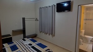 Quarto com cama, ventilador de teto, televisão e banheiro visível por uma porta aberta.