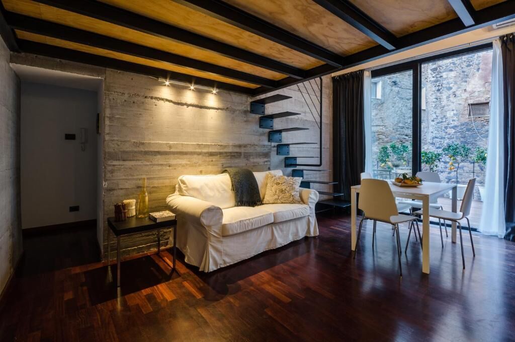 Uma sala de estar moderna com teto de madeira, um sofá branco, uma mesa de jantar com cadeiras e vista para uma parede de pedra e vegetação lá fora.