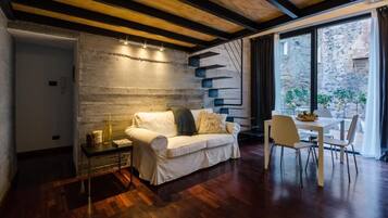 Uma sala de estar moderna com teto de madeira, um sofá branco, uma mesa de jantar com cadeiras e vista para uma parede de pedra e vegetação lá fora.
