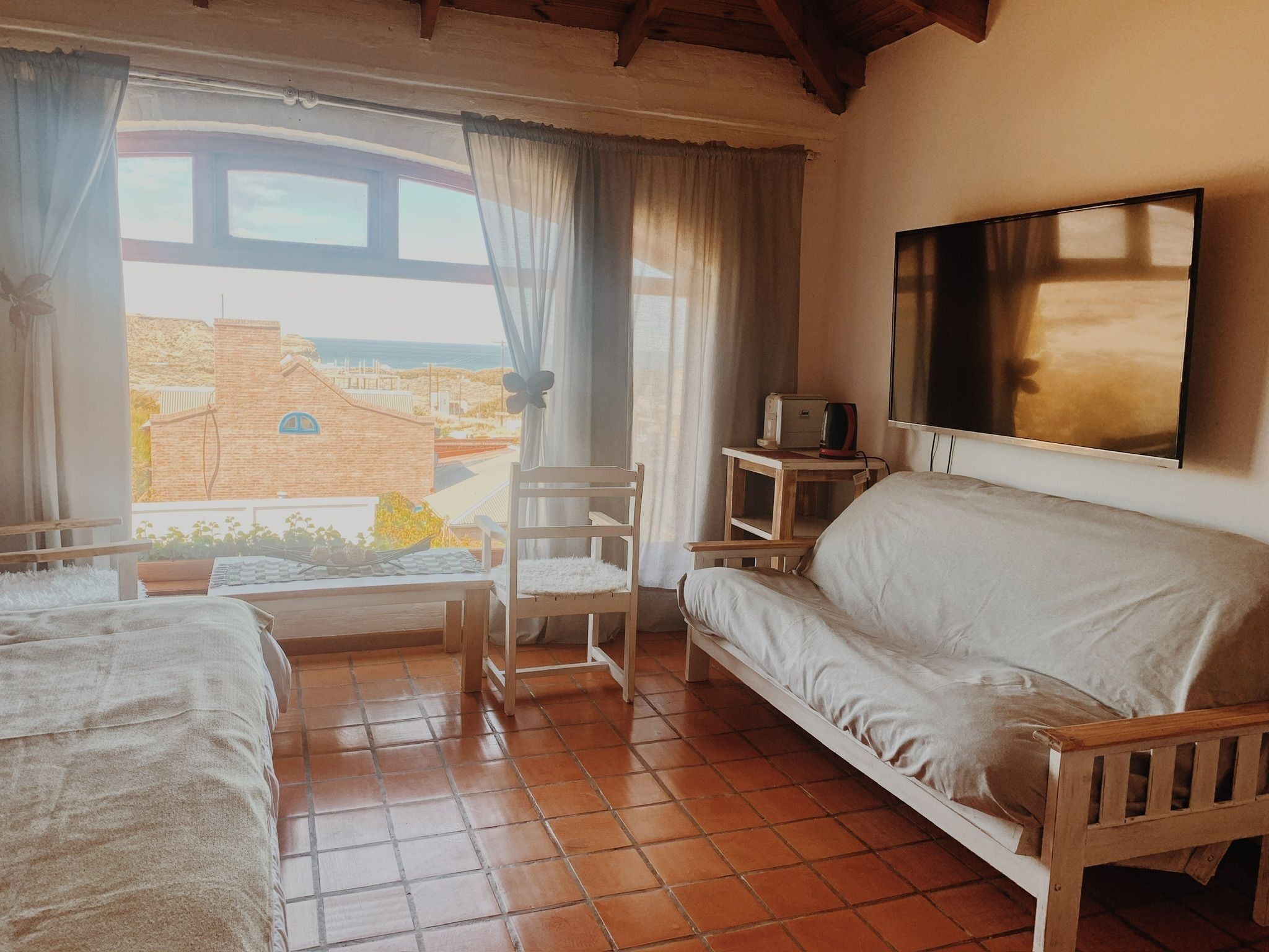 Quarto com cama, cadeira de madeira, sofá, televisão e vista para edifícios pela janela.