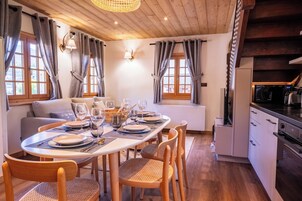 Uma área de refeições com uma mesa redonda posta para quatro pessoas, cadeiras de madeira, um sofá e uma cozinha com frigorífico e fogão.