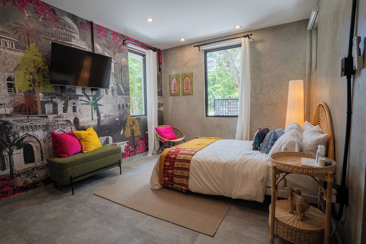 Quarto com cama, sofá verde, TV na parede e janela com vista para árvores.