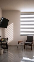 Uma sala de estar moderna com televisão de tela plana na parede, uma mesa de jantar com cadeiras, um banquinho pequeno e uma janela com persianas.