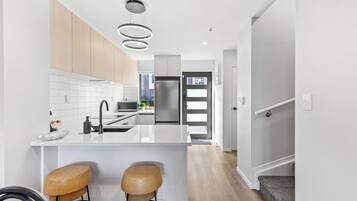 Uma cozinha moderna com armários brancos, uma ilha central com banquetas, eletrodomésticos de aço inoxidável e um lustre pendente.