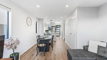 Uma sala de estar moderna com um sofá cinzento, um espaço de refeições com mesa e cadeiras, e uma cozinha ao fundo.