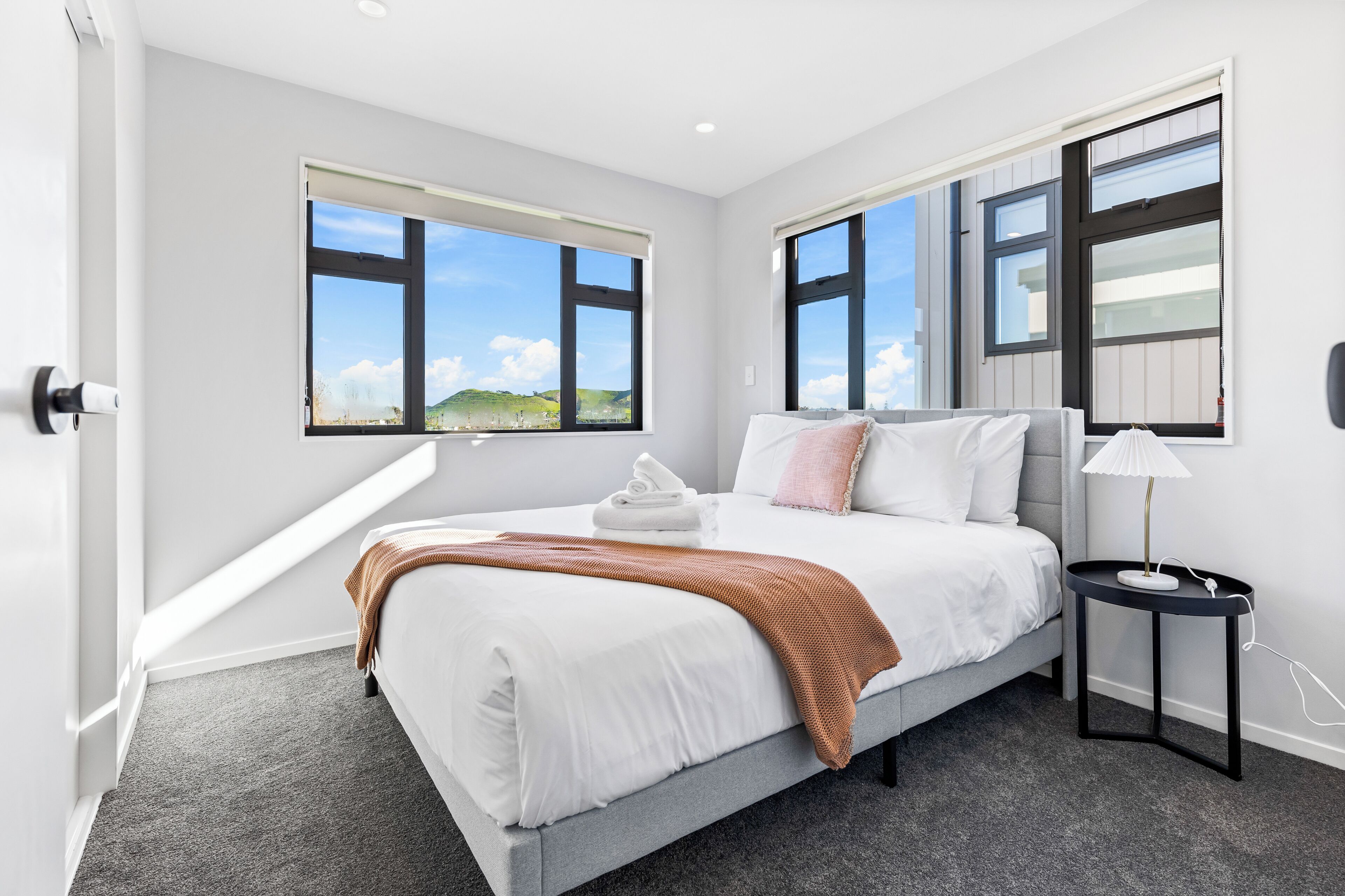 Quarto moderno com uma cama grande, uma mesinha de cabeceira e vista para o exterior através de duas janelas.
