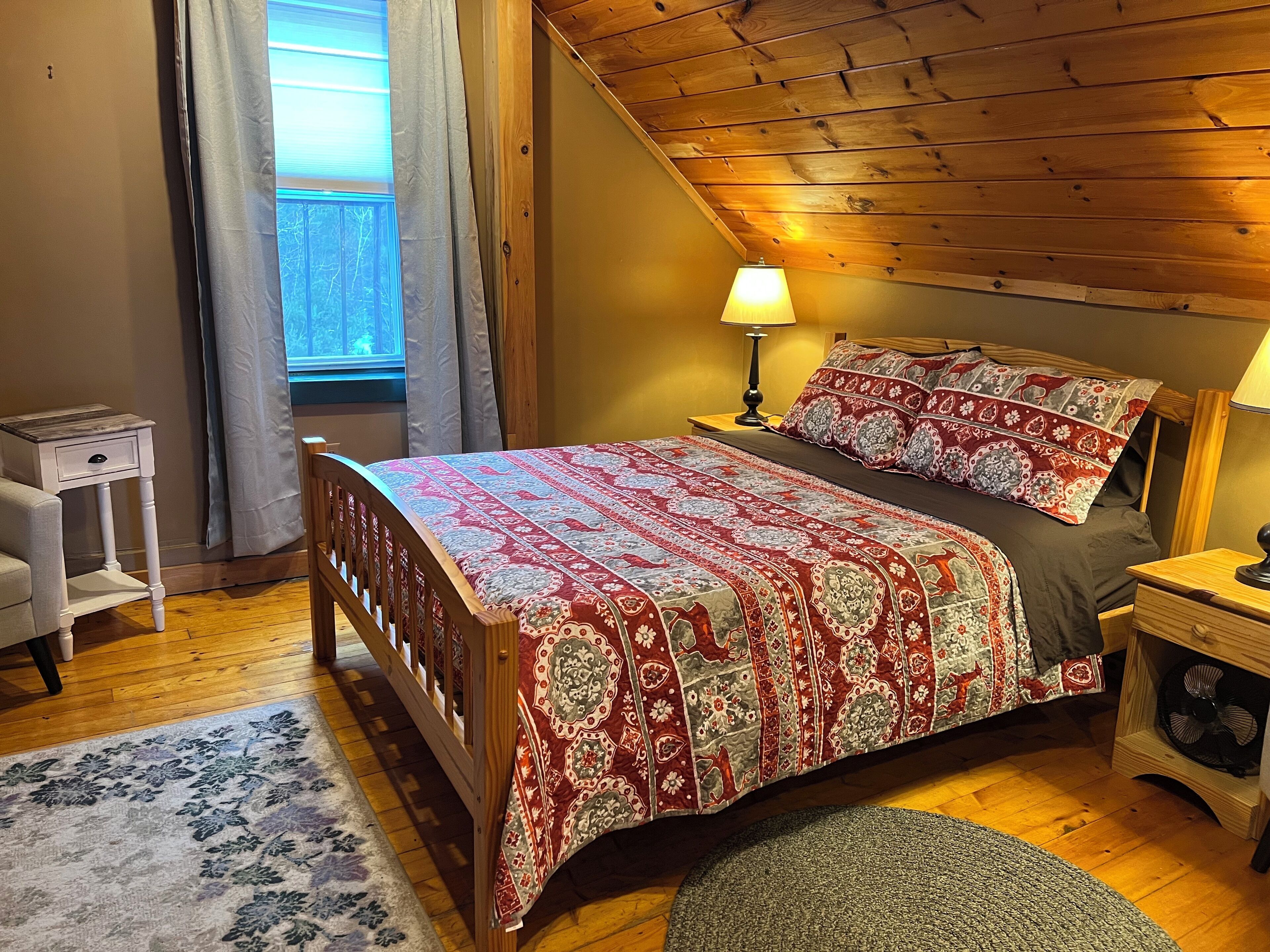 Quarto com teto de madeira, cama com colcha estampada, mesa de cabeceira com abajur, uma cadeira e janela com cortinas.