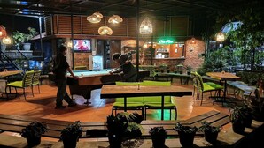 Uma área de refeições ao ar livre com mesas de madeira e assentos acolchoados em verde, cercada por plantas em vasos.