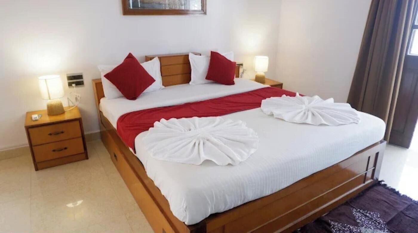 Cama bem arrumada com estrutura de madeira, roupa de cama branca e almofadas vermelhas. Há duas mesas de cabeceira com luminárias, uma mesinha de apoio de madeira e uma janela com cortinas.