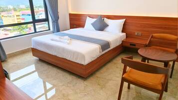 Quarto de hotel com uma cama grande, cabeceira de madeira, uma mesa pequena, uma cadeira e vista para prédios pela janela.