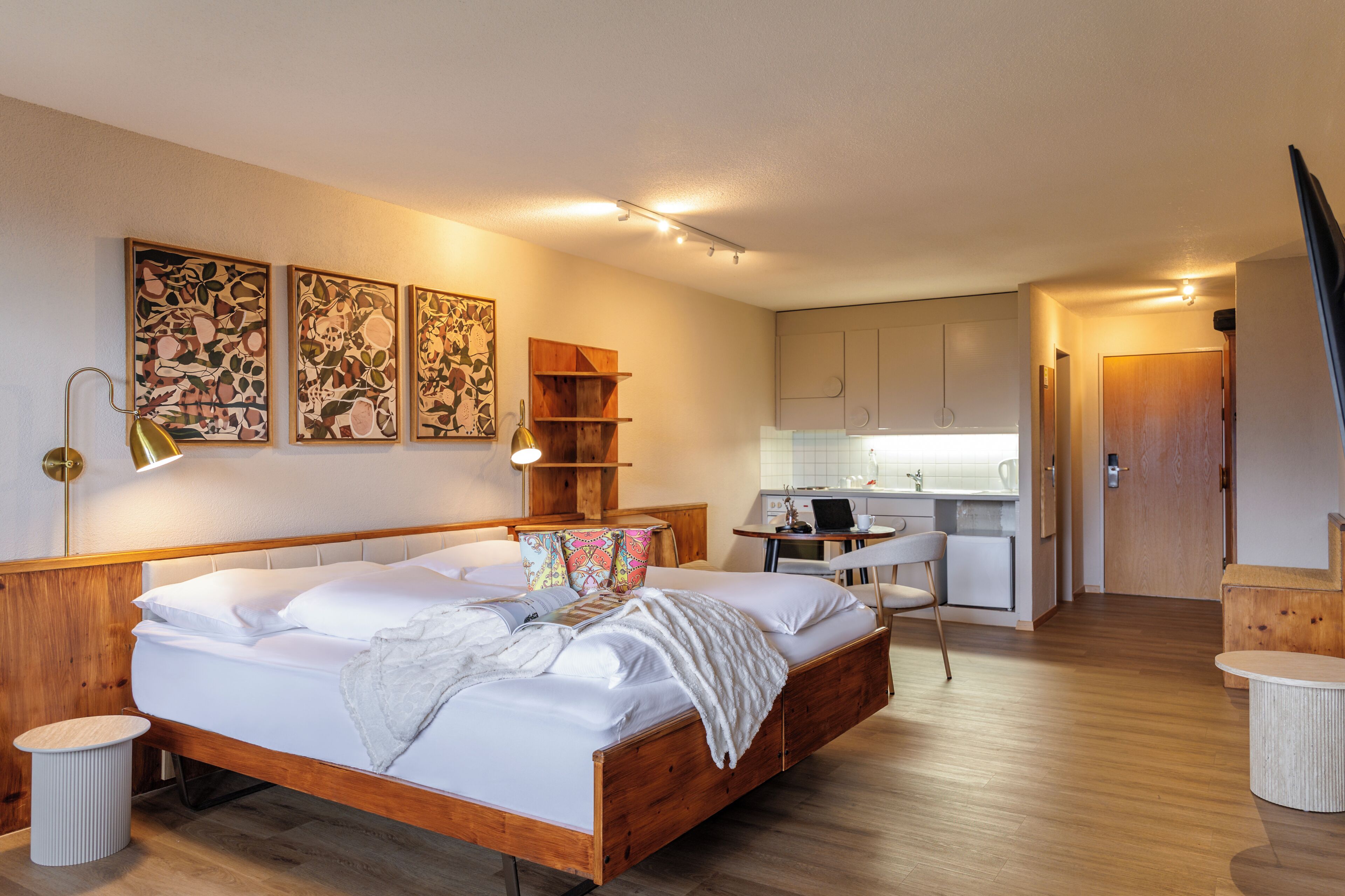Quarto moderno com cama de madeira, mesas de cabeceira e escrivaninha com cadeira. Possui também uma cozinha com armários e pia.