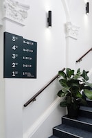 Escada com corrimão preto, um vaso de planta e uma placa na parede indicando números de quartos e seus respectivos tipos.