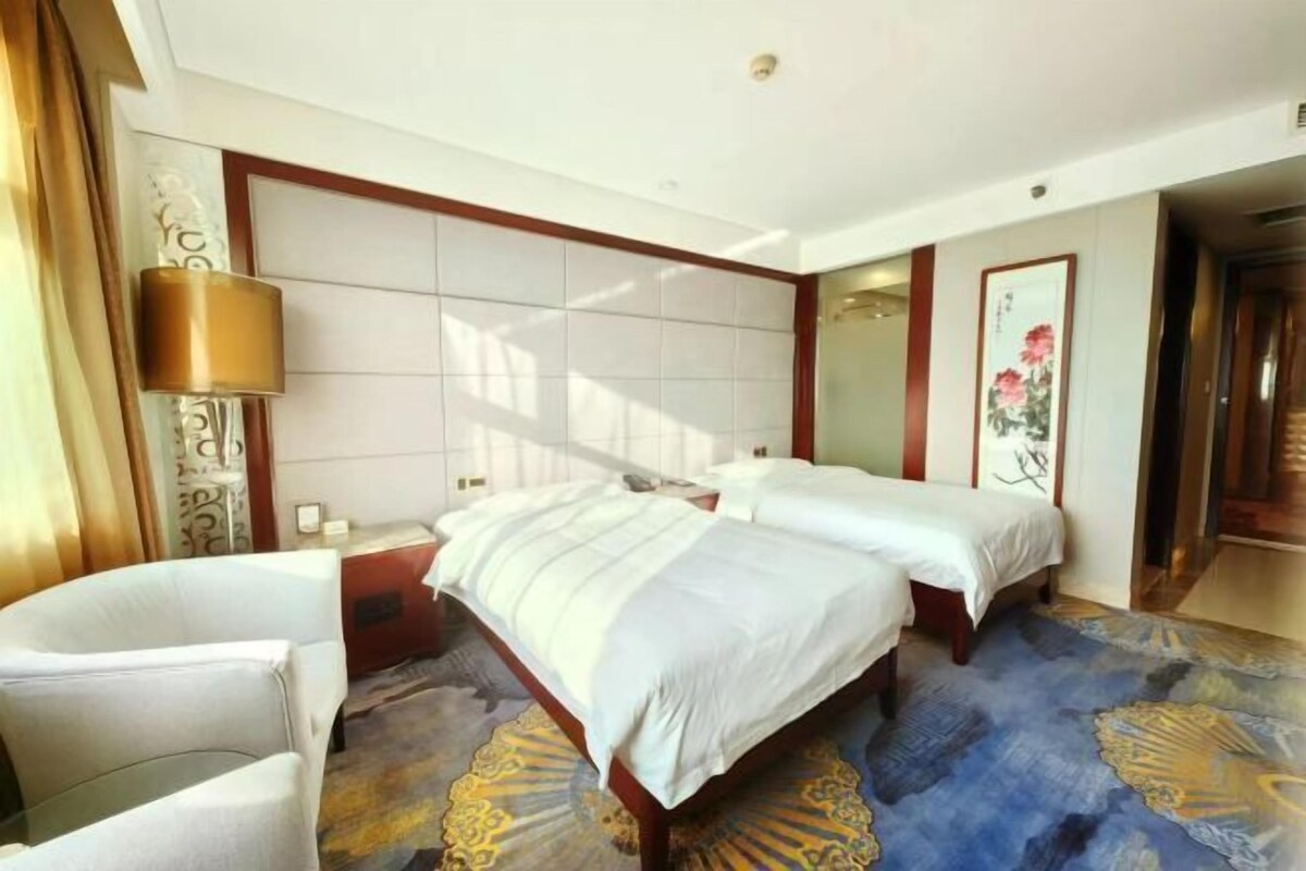 Quarto de hotel com duas camas, um espaço de estar com uma poltrona, uma janela grande e um painel decorativo na parede.
