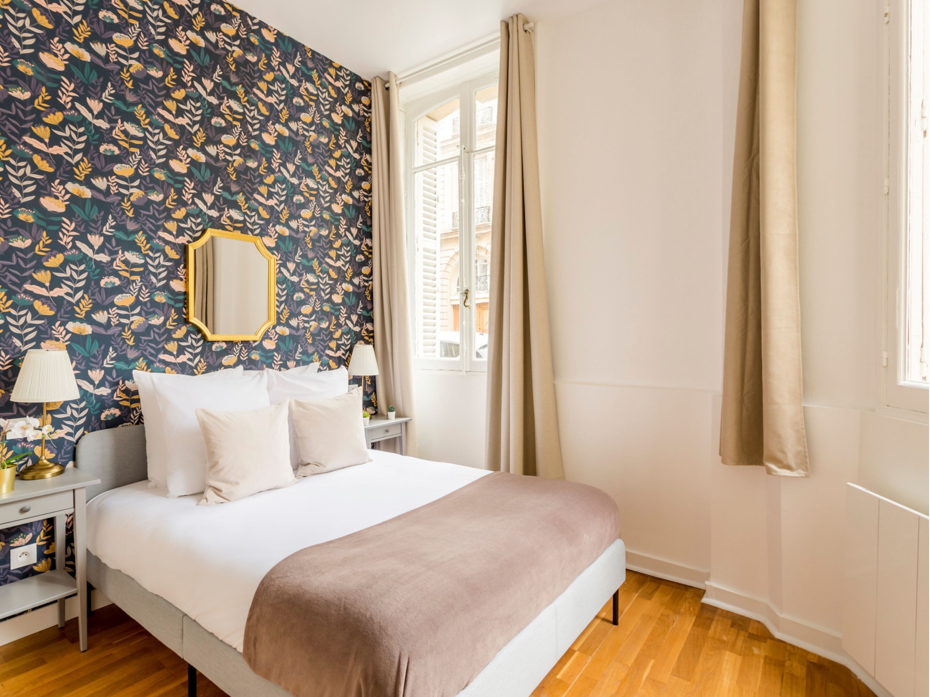 Quarto bem arrumado com papel de parede floral, uma cama grande com roupa de cama branca, uma mesa de cabeceira com abajur e uma janela com cortinas.