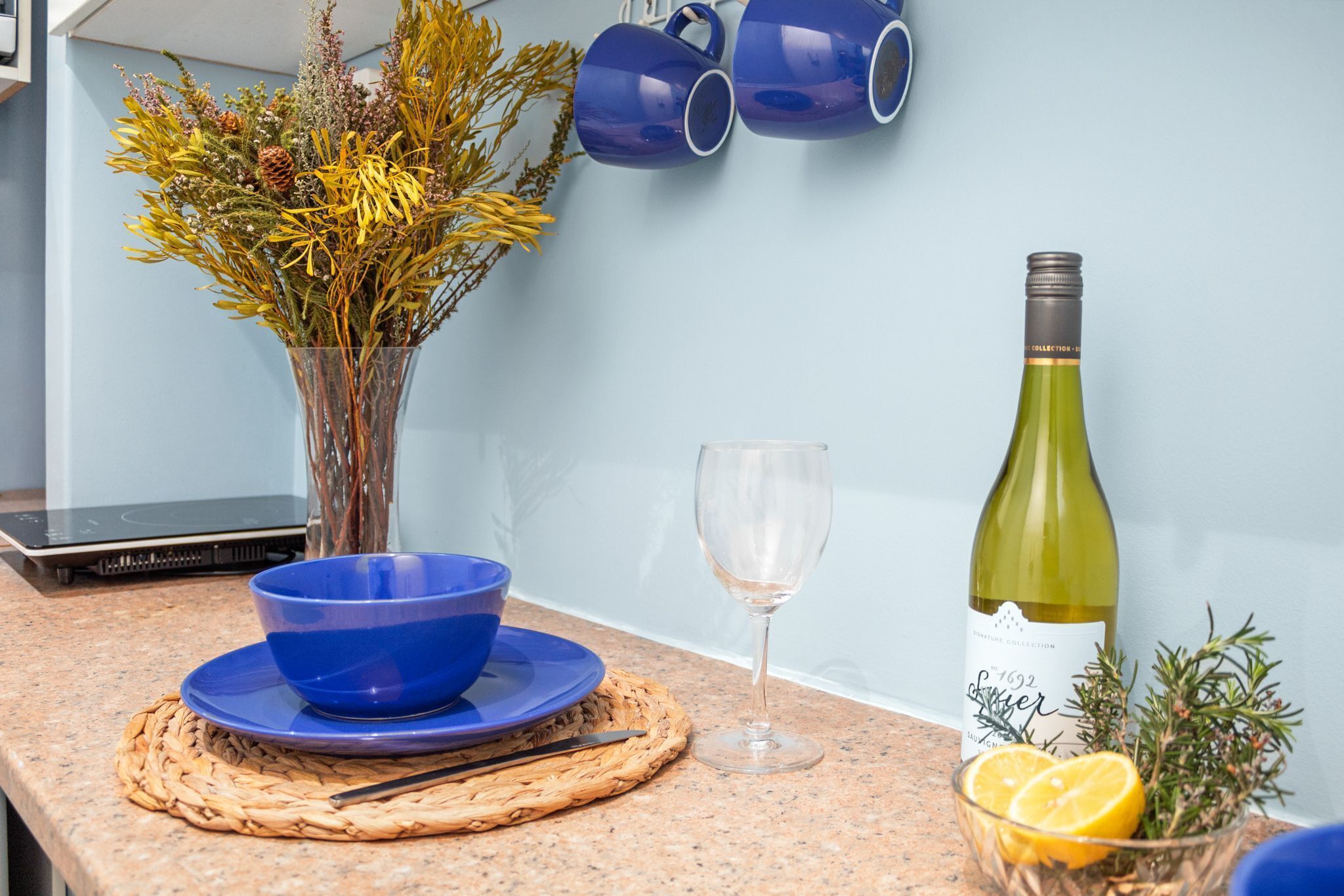 Uma bancada de cozinha com uma tigela azul, uma taça de vinho, uma garrafa de vinho e um vaso com flores secas.
