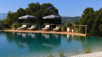 Piscina com água cristalina, rodeada por espreguiçadeiras e guarda-sóis, com árvores e colinas ao fundo.