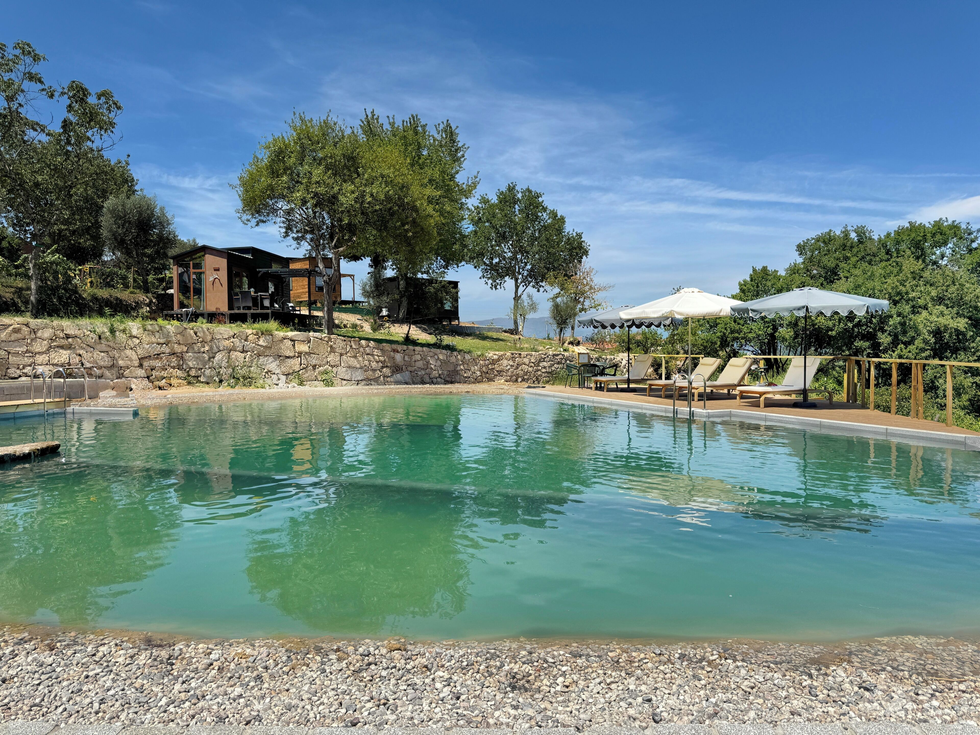 Uma piscina com espreguiçadeiras e guarda-sóis, cercada por um muro de pedra e árvores.