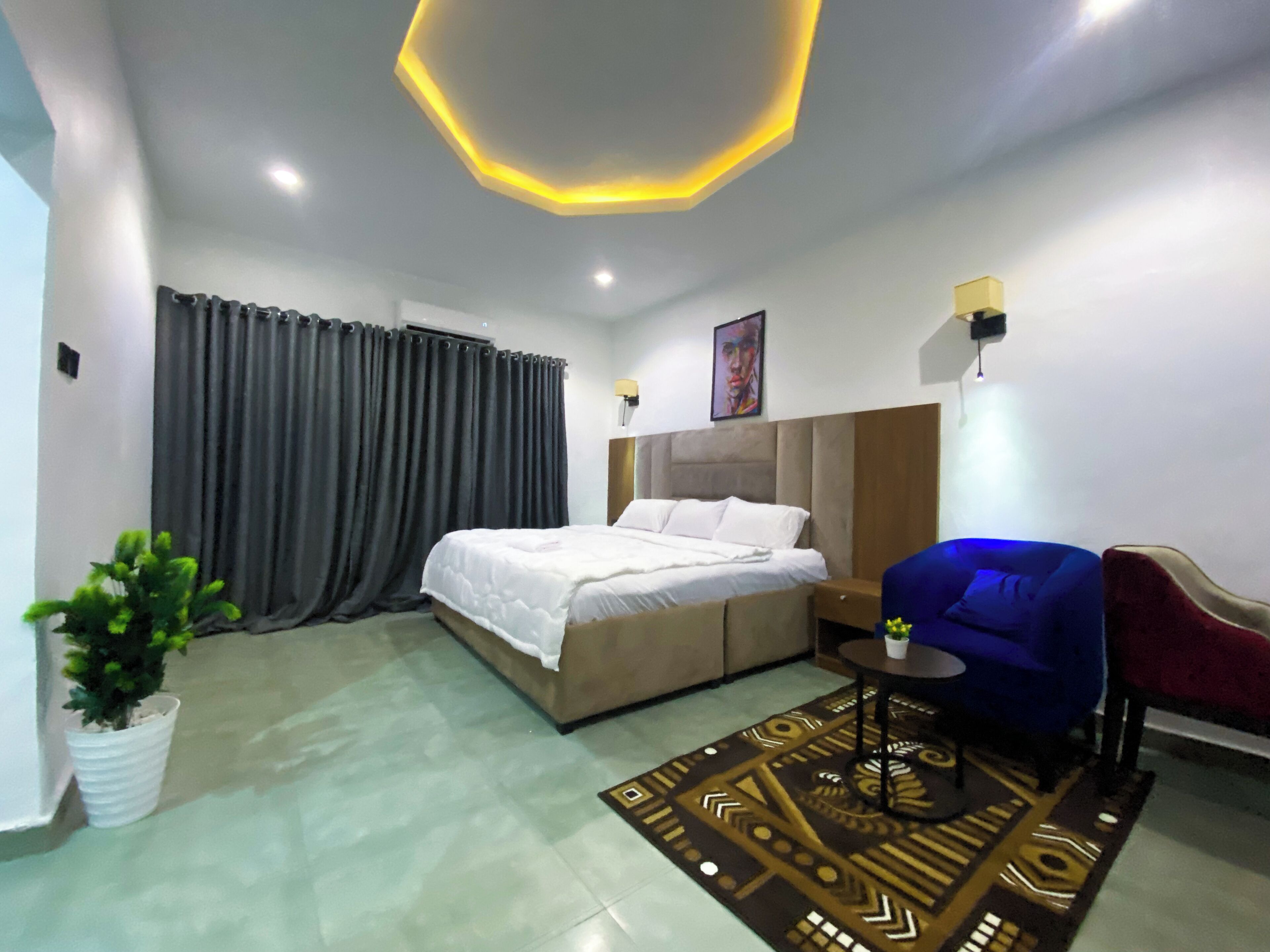 Quarto de hotel moderno com uma cama grande, uma poltrona azul, uma mesinha e um tapete decorativo.