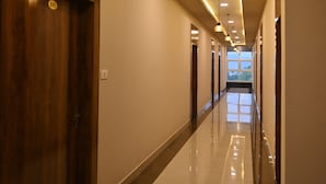 Corredor longo e bem iluminado, com portas de madeira e piso brilhante, que leva a um quarto claro com uma janela grande.