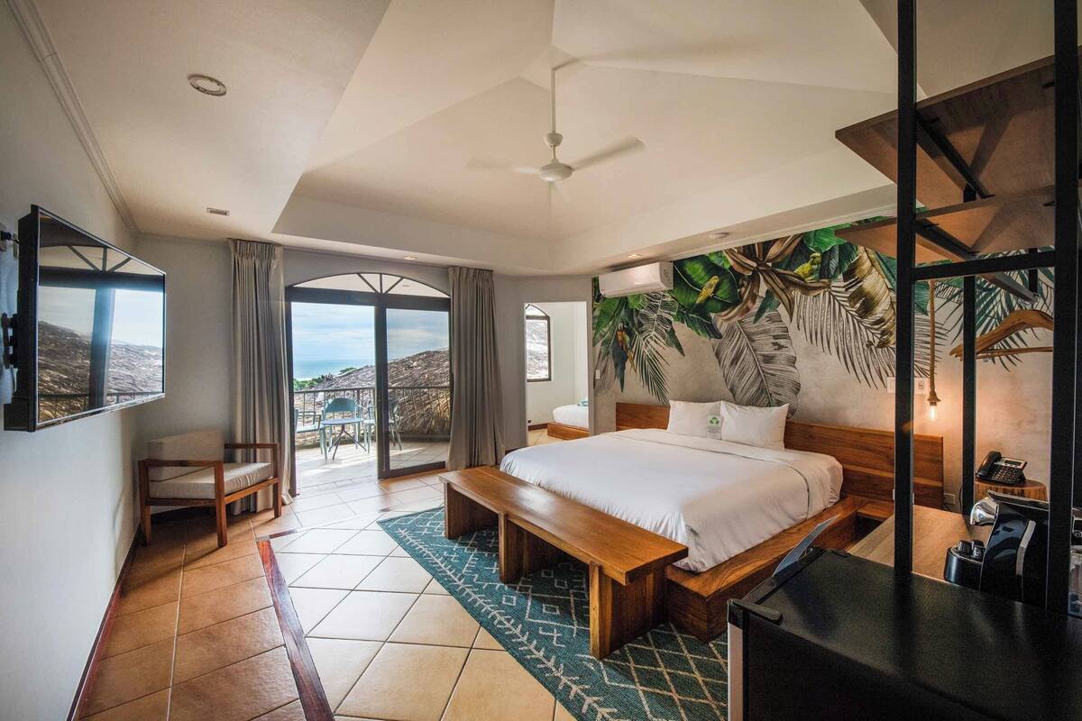 Quarto espaçoso com uma cama grande, um banco de madeira, uma escrivaninha e vista através de uma porta de vidro.