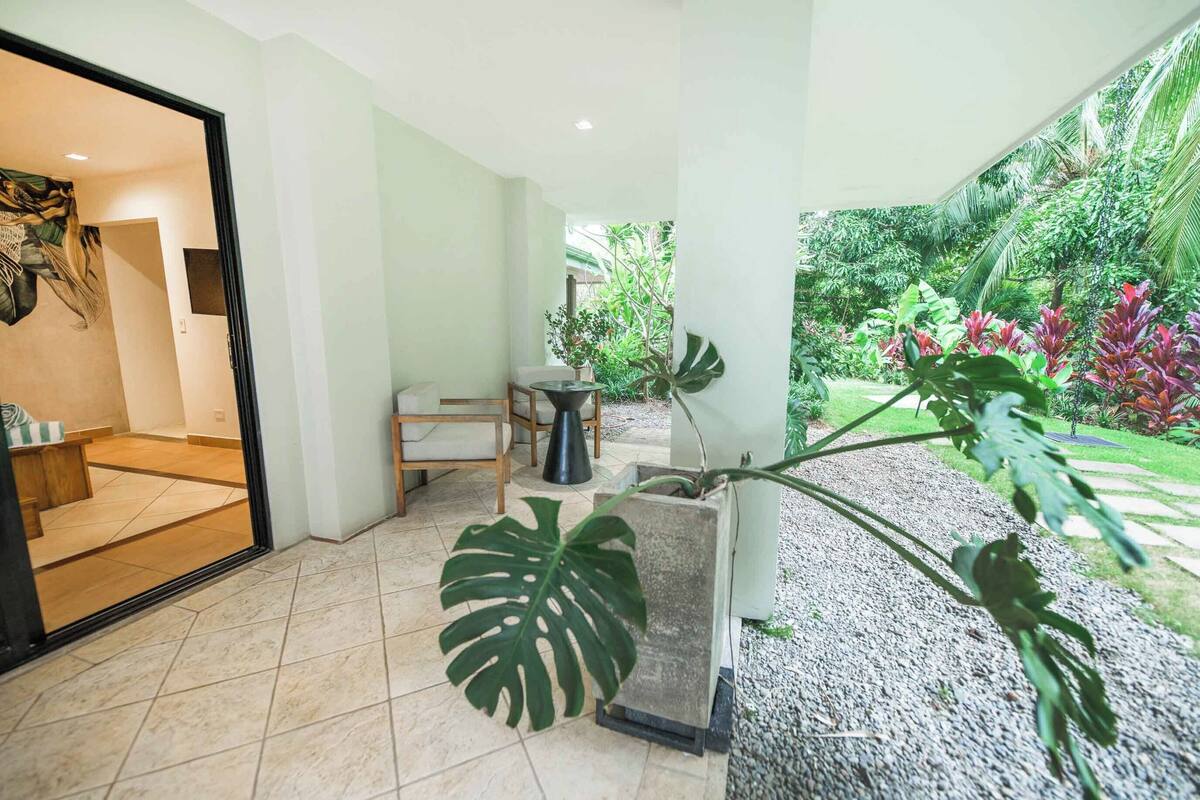 Um interior moderno com piso ladrilhado, área de estar com mesa e cadeiras, e vista para um jardim exuberante com diversas plantas.