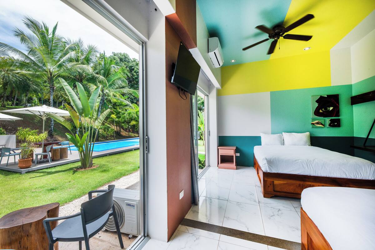 Quarto com cama grande, ventilador de teto e vista para a piscina e o jardim.